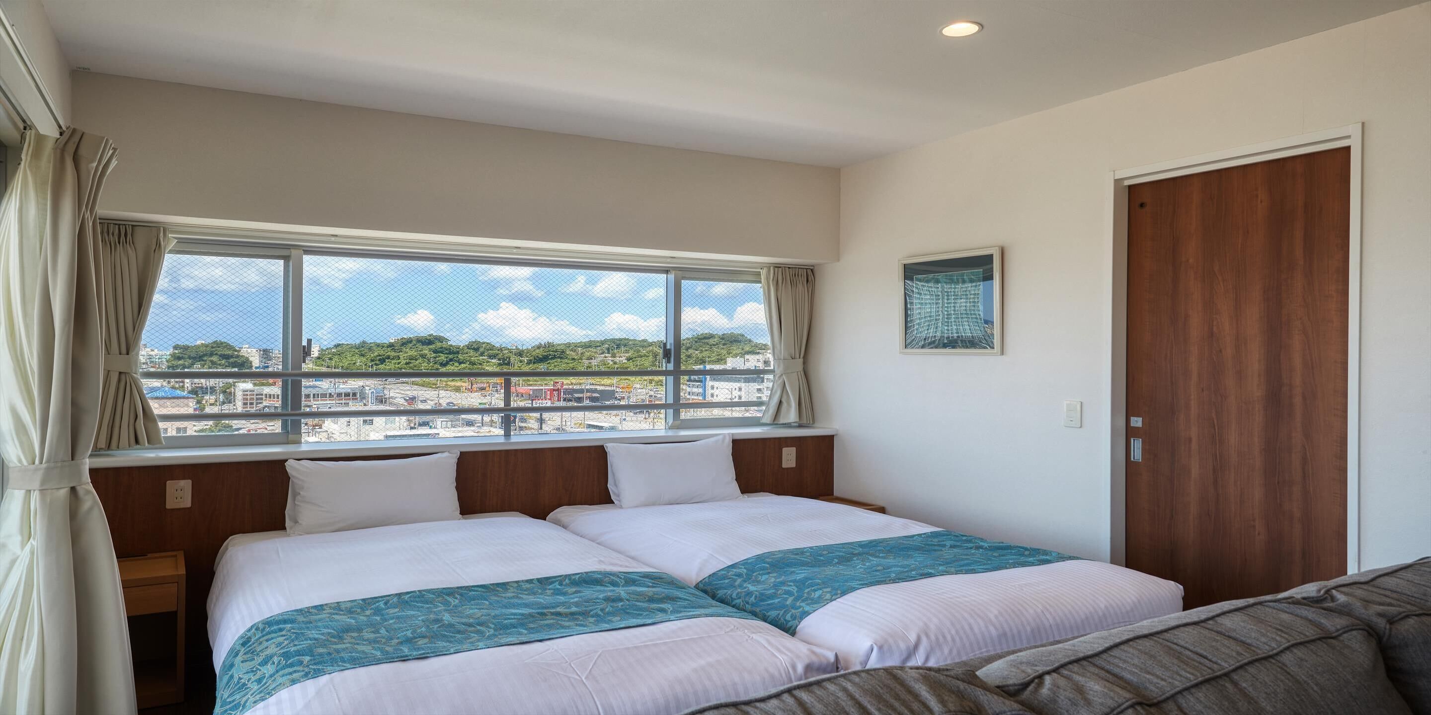 Quarto de hotel com duas camas, uma janela grande com vista para a cidade, uma porta de madeira e uma pequena imagem na parede.