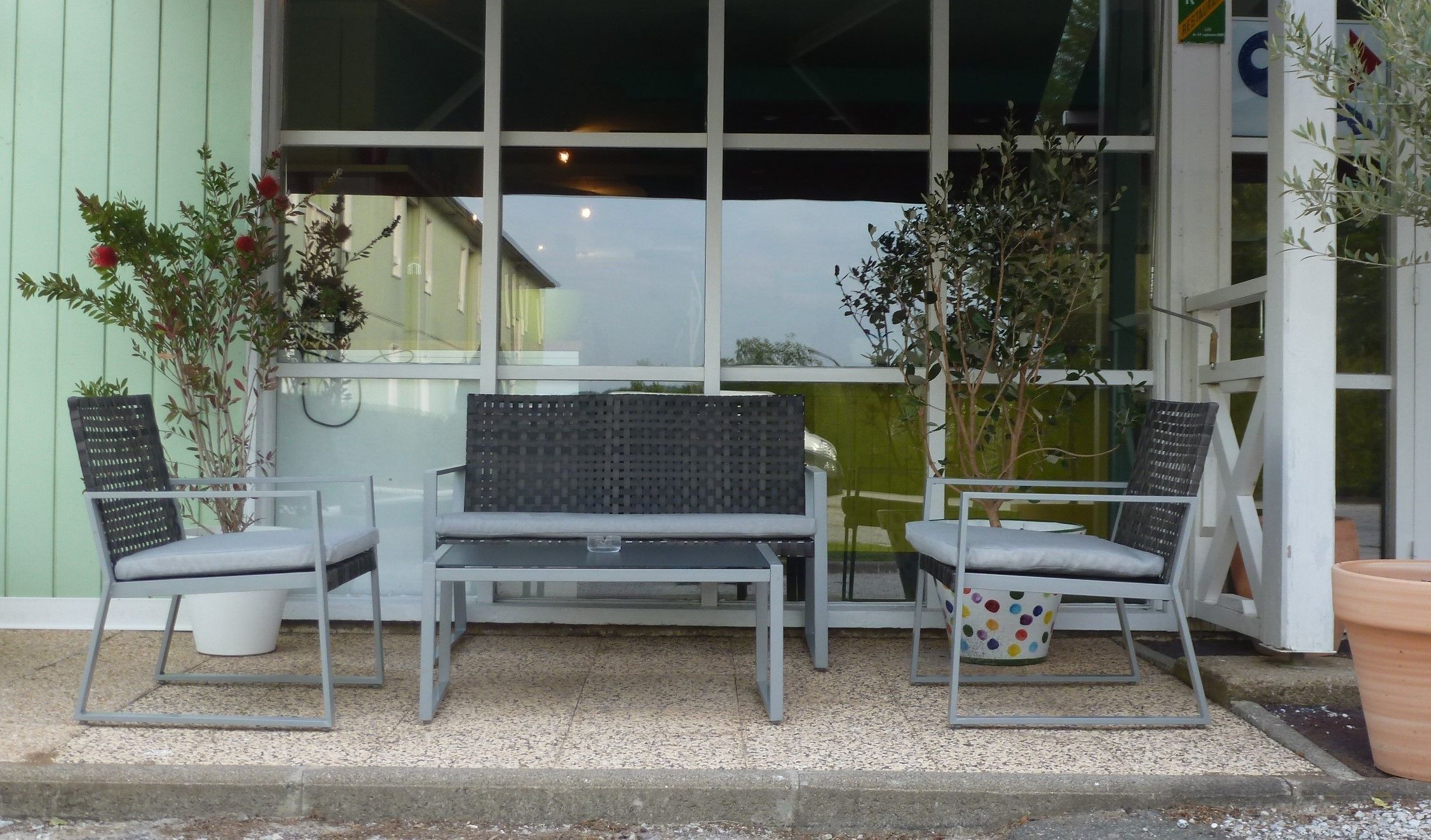 Pátio com duas cadeiras, uma mesa, planta em vaso e janela com vista para um edifício.