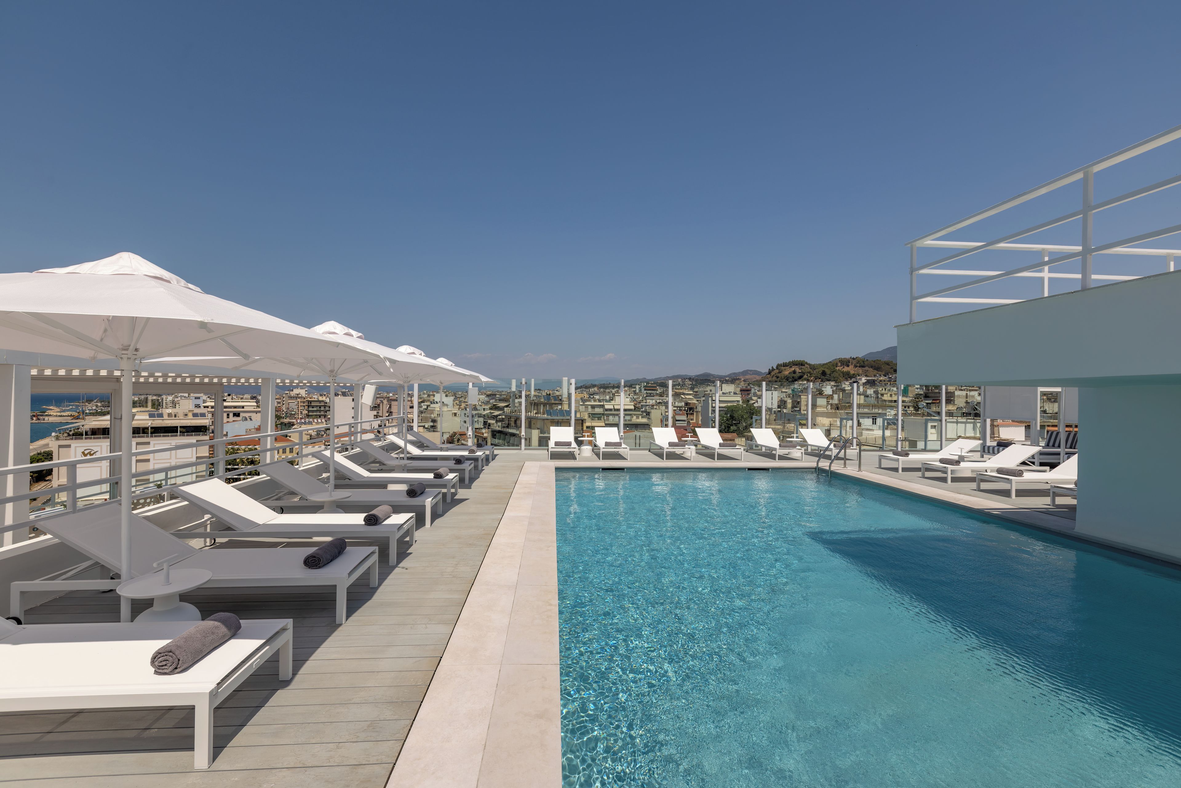 Área de piscina no terraço com espreguiçadeiras e guarda-sóis, com vista para a paisagem urbana.