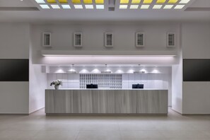Bancada de recepção moderna com acabamento em madeira clara, um vaso de planta e uma grade de luzes acima.