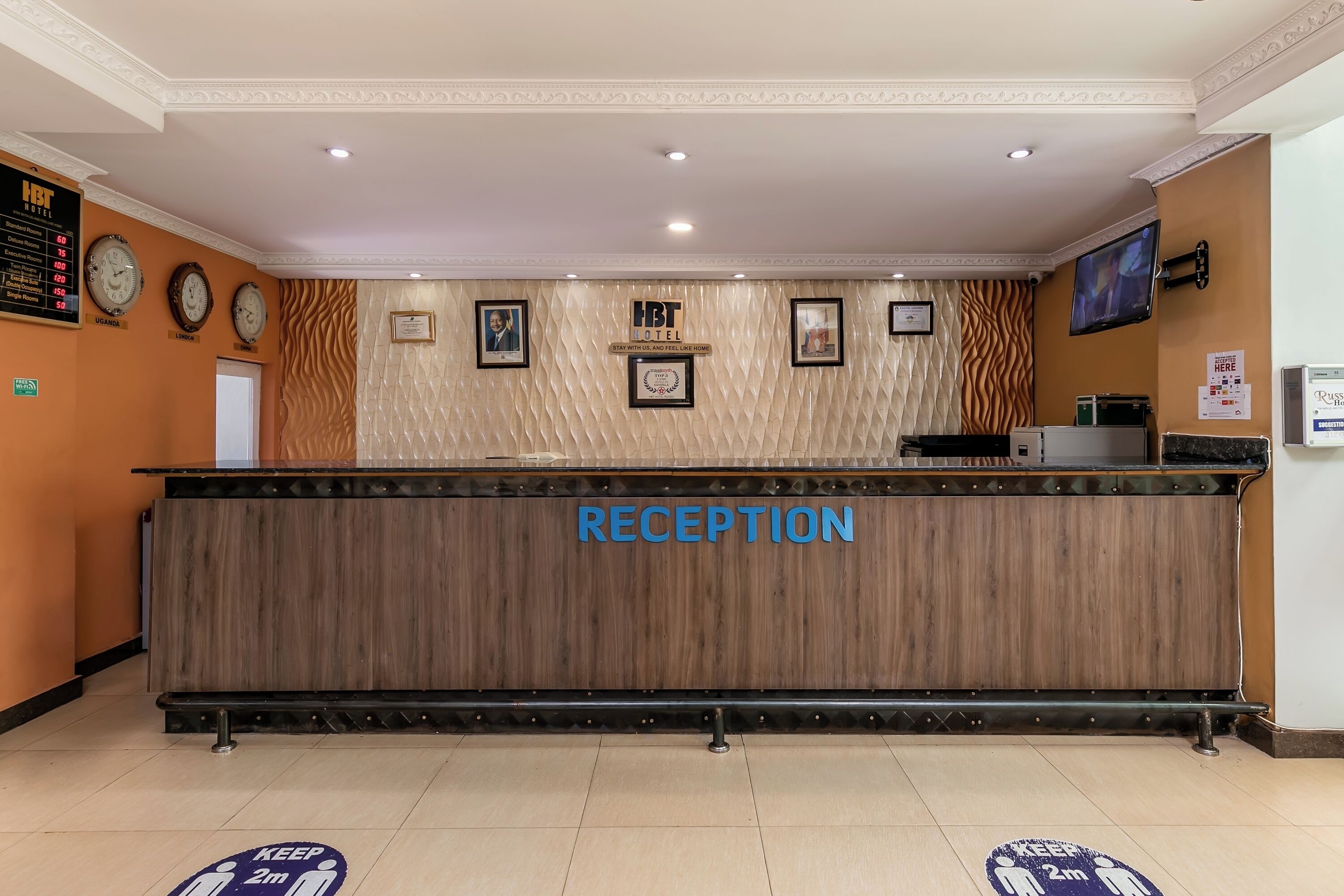 Uma área de recepção de hotel com um balcão de madeira identificado como “RECEPTION”, uma TV na parede e quadros emoldurados.