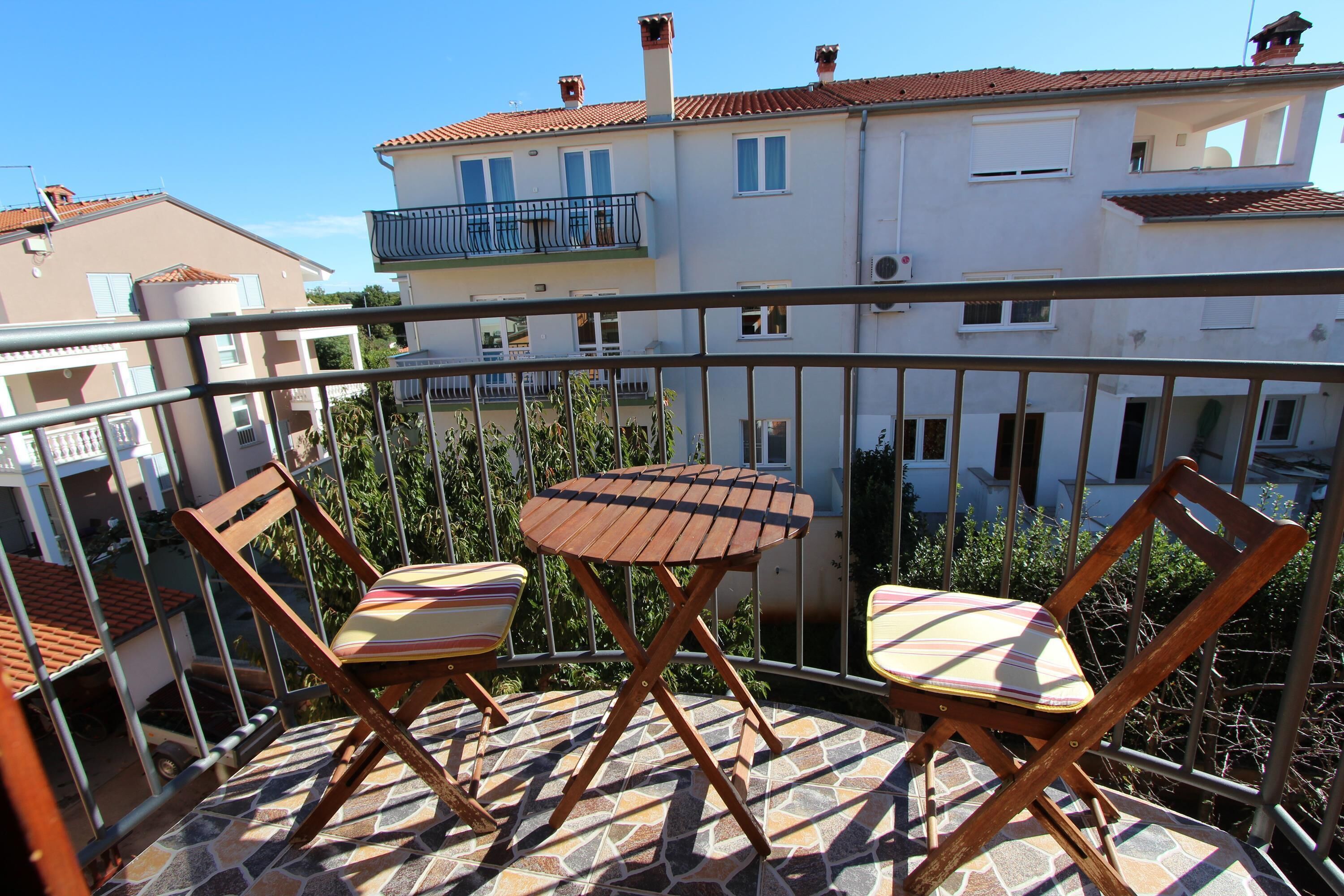Varanda com duas cadeiras de madeira e uma mesa redonda pequena, com vista para uma área residencial com edifícios e céu claro.