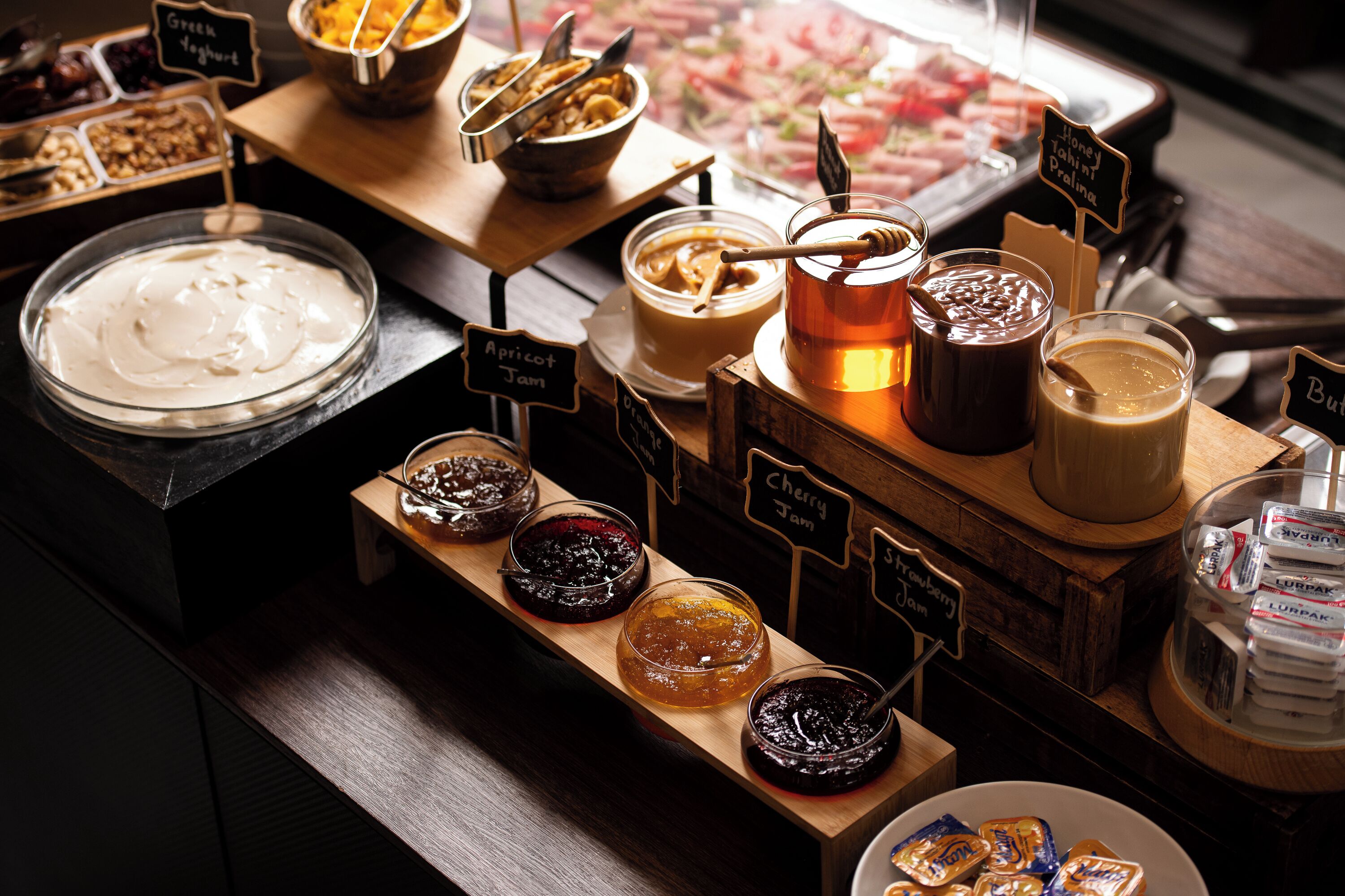 Buffet com diversos alimentos e acompanhamentos, incluindo iogurte grego, iogurte com mel e diferentes geleias.