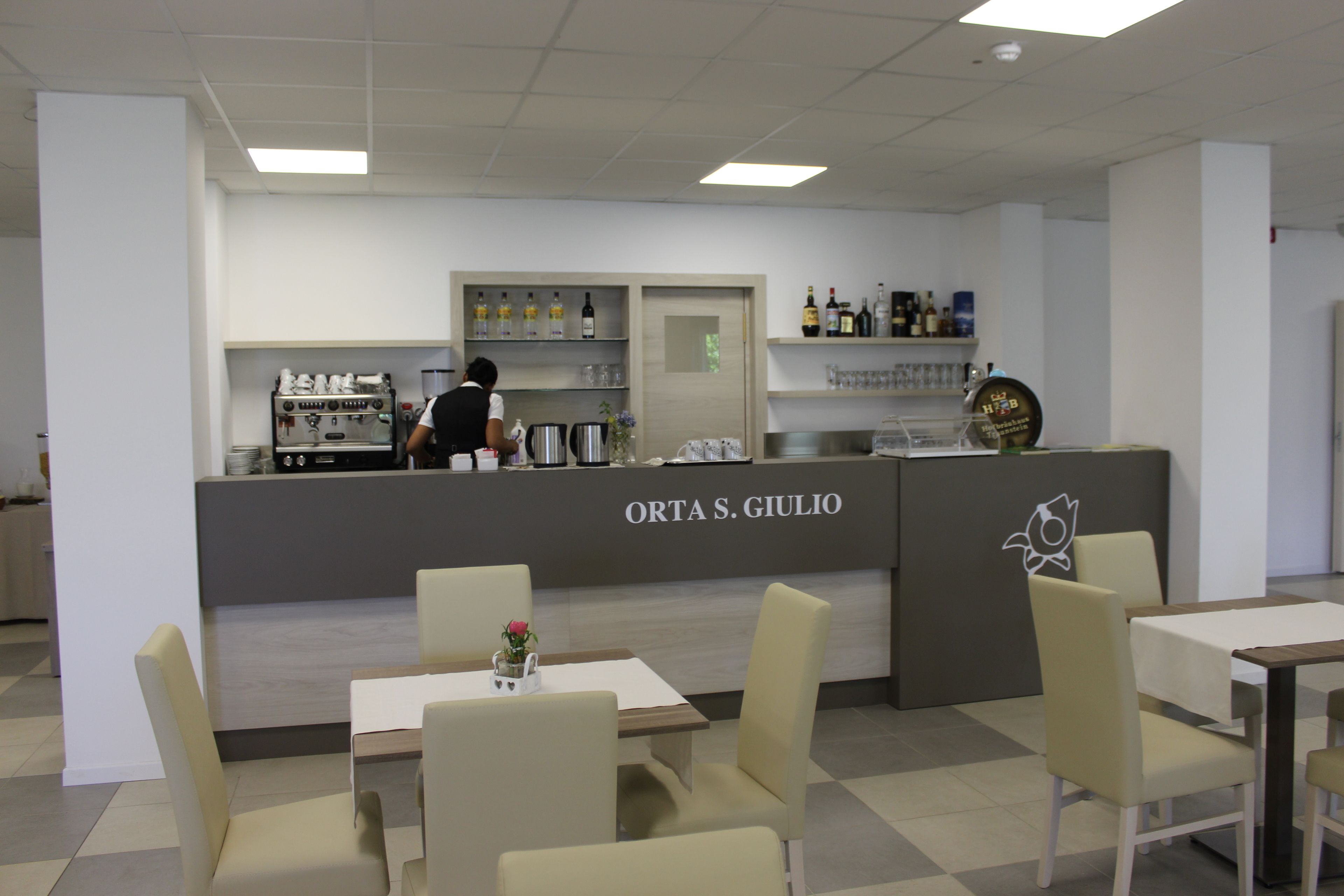 Interior de restaurante com balcão chamado 'ORTA S. GIULIO', área de estar e bar com diversos frascos.