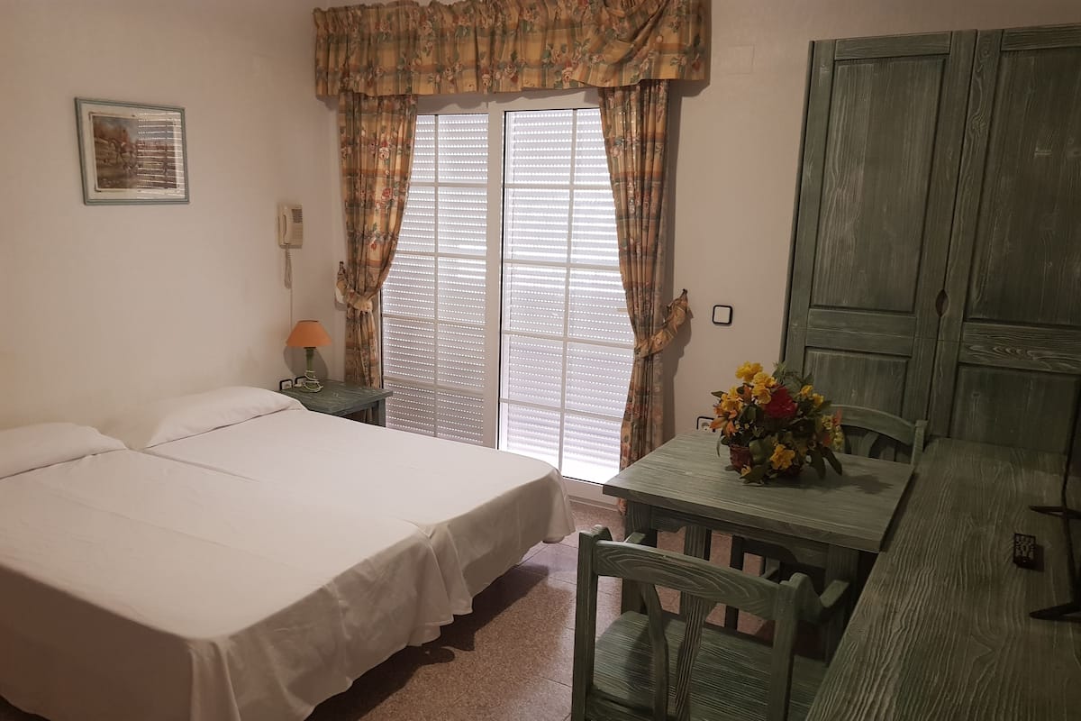 Quarto com cama, mesa de cabeceira, abajur, janela com cortinas e uma mesa com um vaso de flores.
