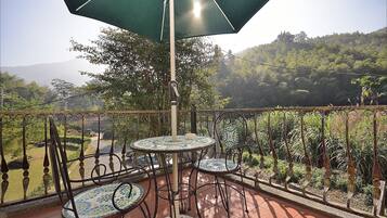 Uma varanda com mesa e cadeiras, um guarda-sol verde e vista para uma paisagem verdejante com colinas ao fundo.