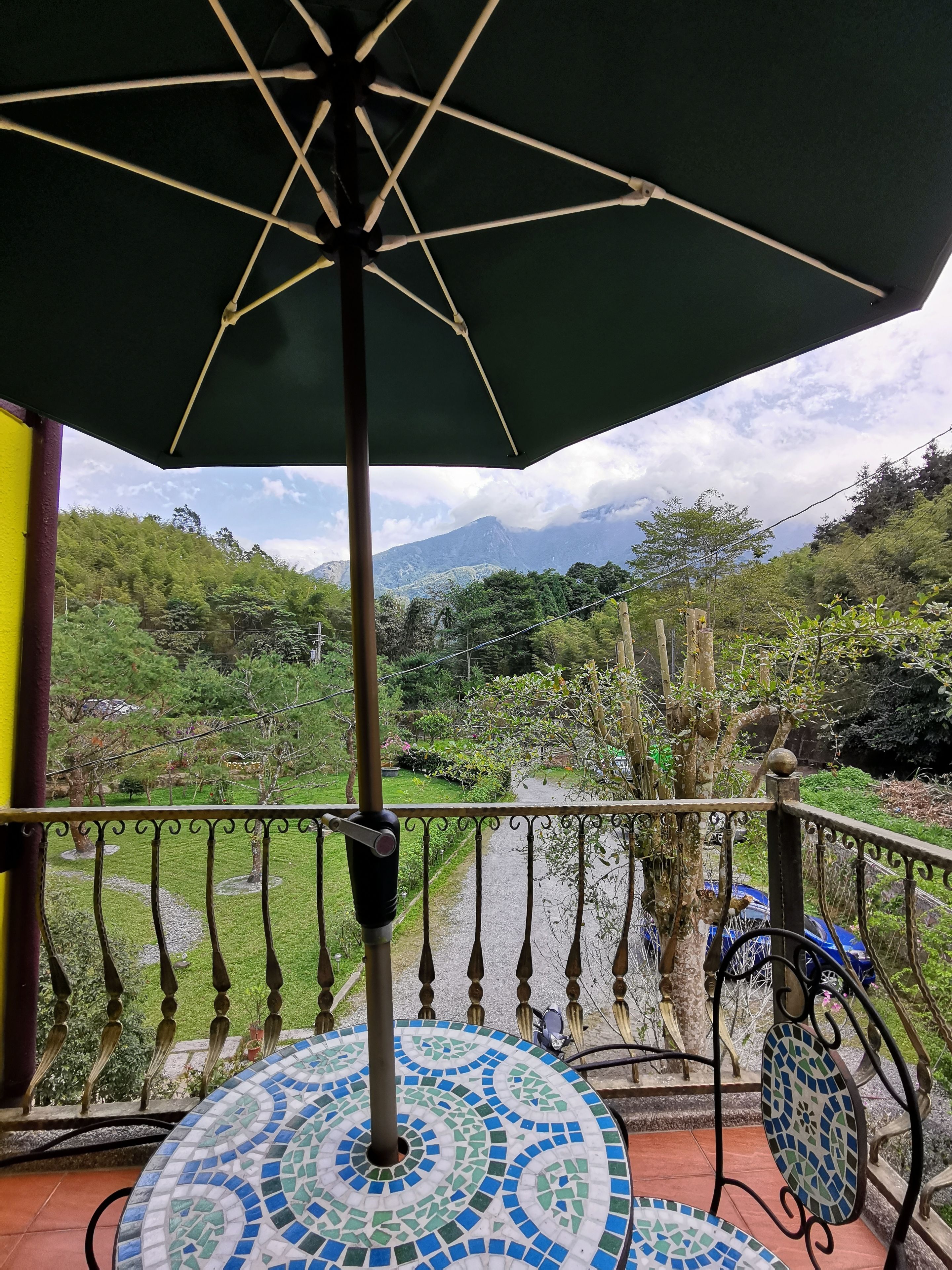 Uma varanda com um guarda-sol verde, uma mesa de azulejos e vista para uma paisagem montanhosa.