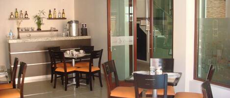 Uma sala de jantar com uma mesa posta para quatro pessoas, rodeada por cadeiras com almofadas laranja. É visível, ao fundo, uma área de cozinha com uma cafeteira e prateleiras cheias de garrafas.