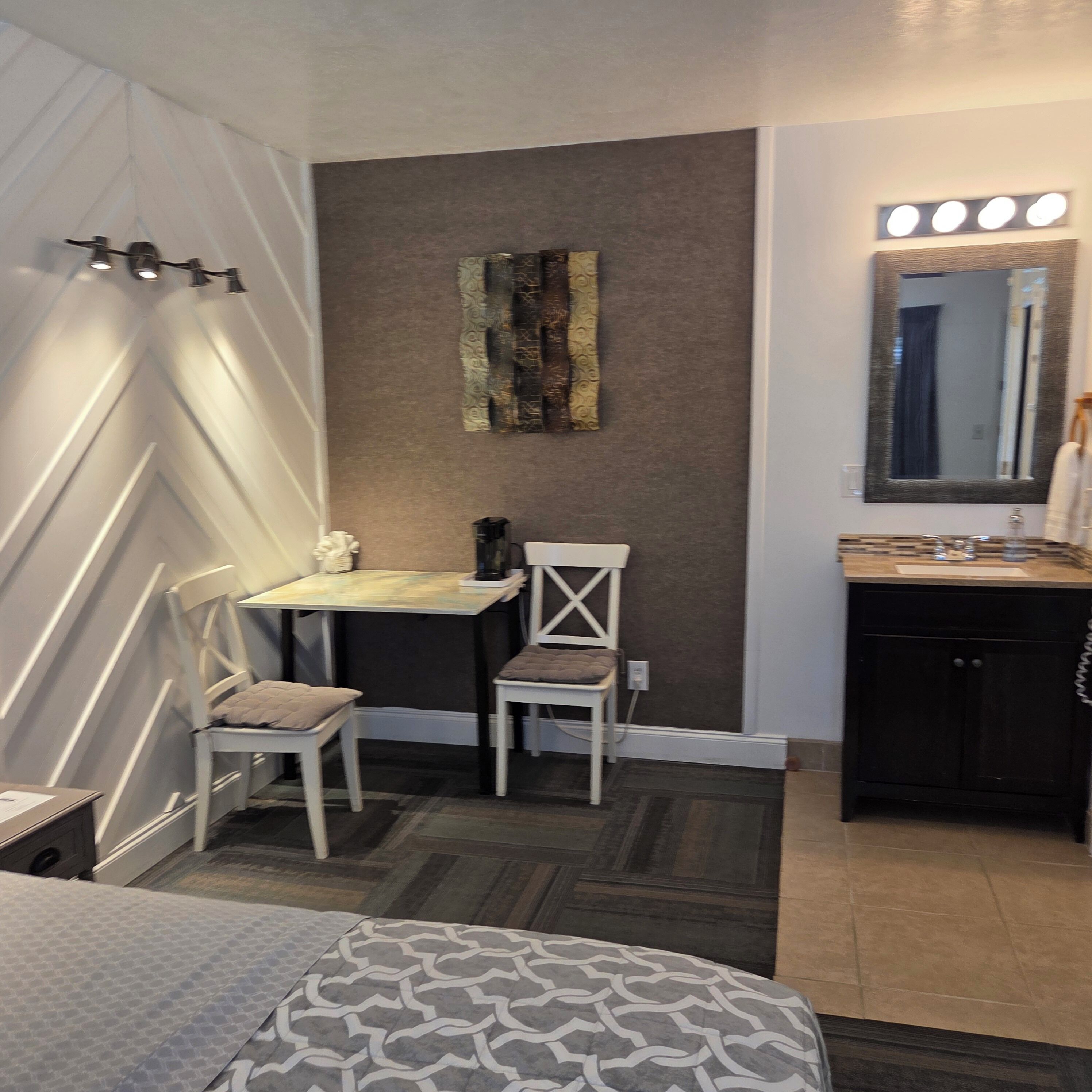 Quarto compacto com cama, uma pequena mesa com cadeira, cafeteira, espelho com iluminação e parede escura.