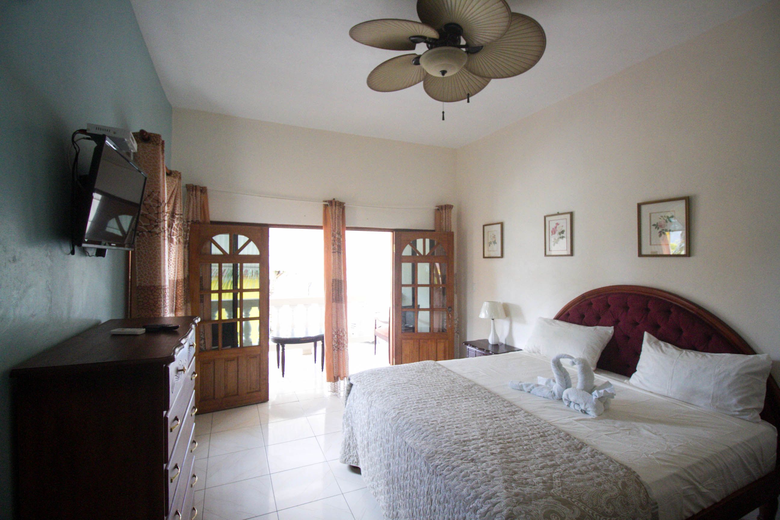 Quarto com cama, cômoda, ventilador de teto, televisão e porta para uma varanda.