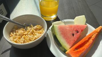 Uma mesa com uma tigela de cereal, um copo de suco de laranja e um prato com fatias de melancia e mamão.