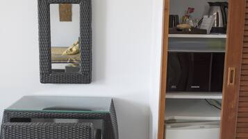 Quarto com espelho trançado na parede, mesa com tampo de vidro e porta de madeira que dá para uma cozinha equipada.