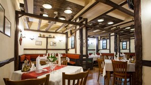 Restaurante com vigas de madeira, toalhas de mesa brancas e assentos acolchoados em vermelho. Mesas dispostas com copos de vinho e talheres.