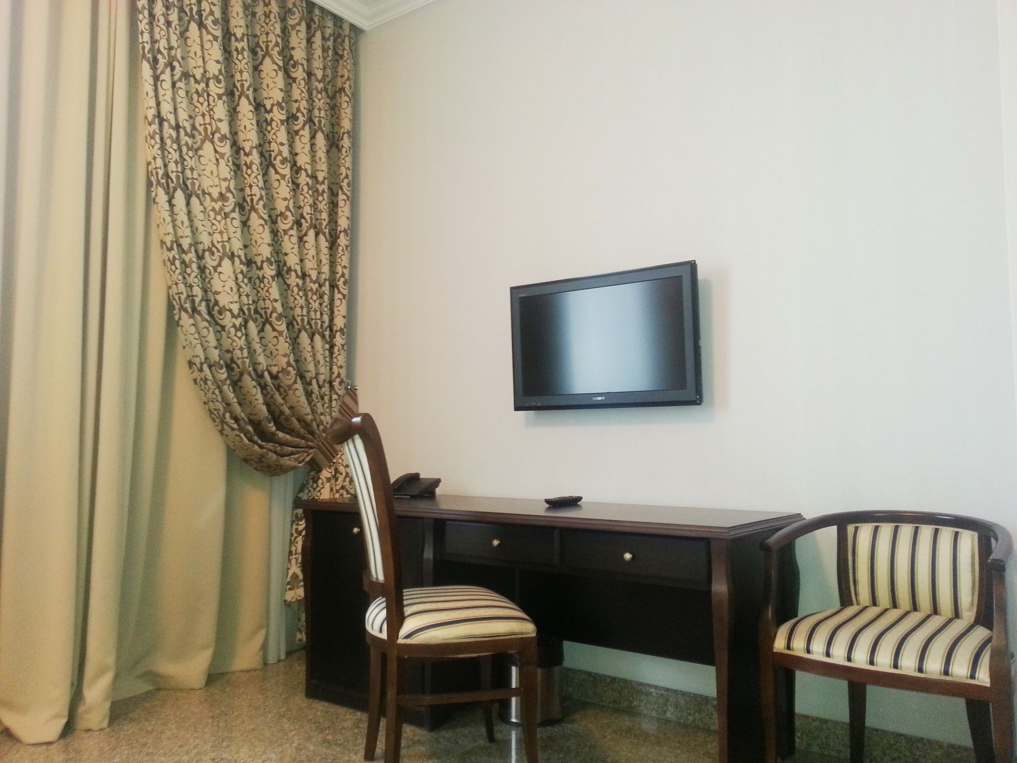 Quarto com escrivaninha, duas cadeiras, televisão e cortina estampada.
