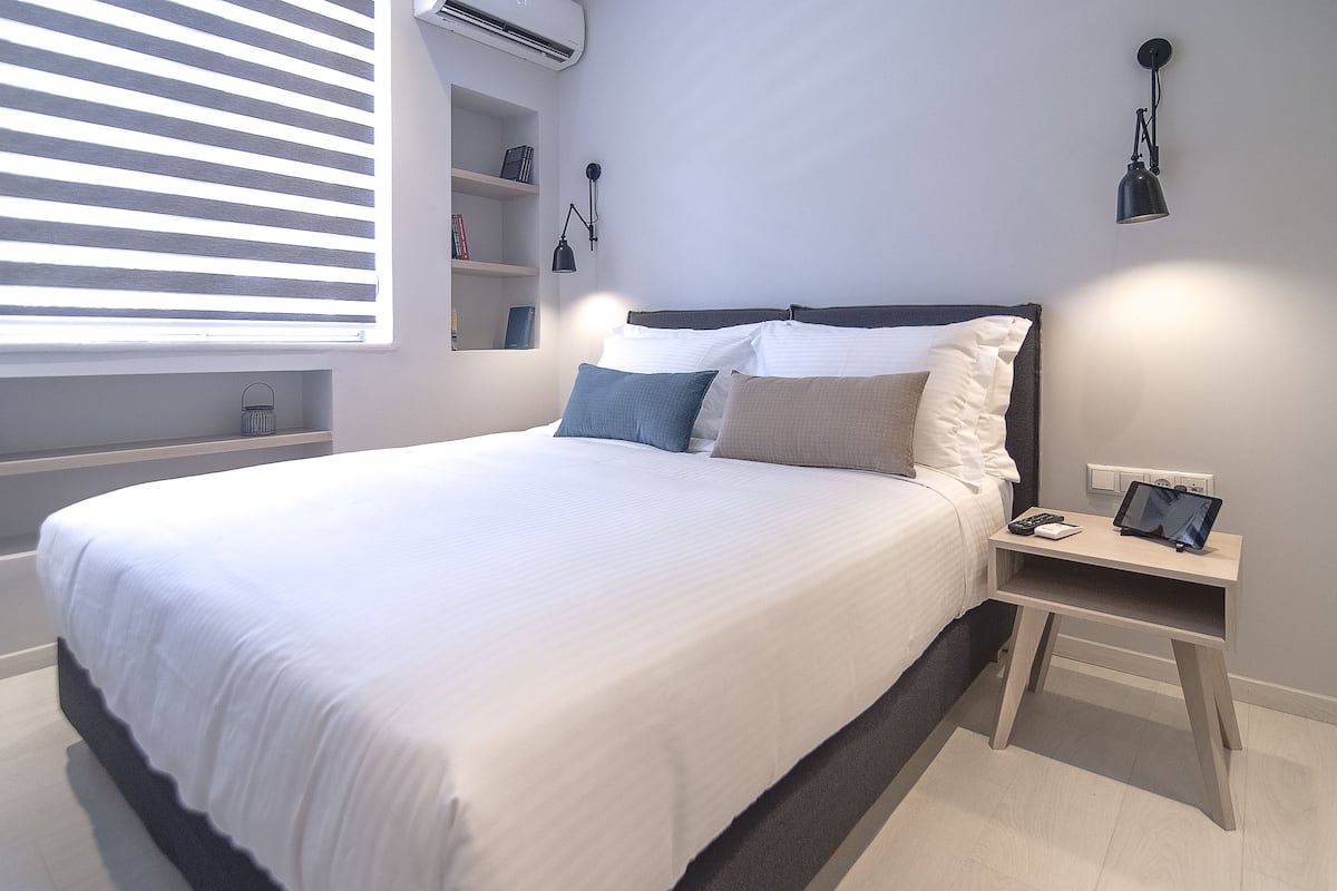 Uma cama bem arrumada com edredom branco e almofadas, uma mesa de cabeceira com abajur, e uma estante com livros e uma pequena planta.