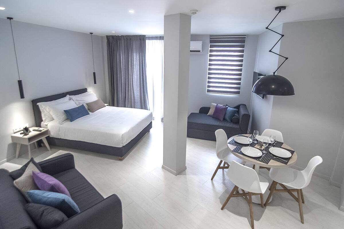 Quarto de hotel moderno com cama, sofá, mesa de jantar com cadeiras e janela com persianas.