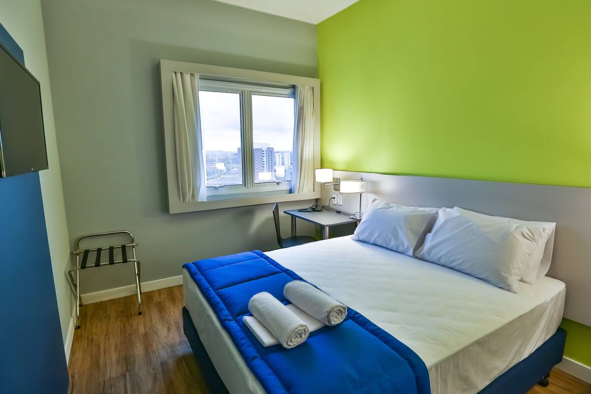 Quarto de hotel com uma cama, janela com cortinas, mesa pequena com abajur e um suporte metálico.