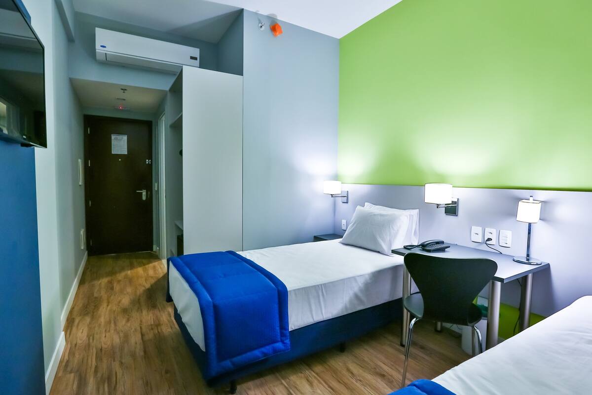 Quarto de hotel com uma cama, uma escrivaninha com cadeira, telefone e luminária na parede.