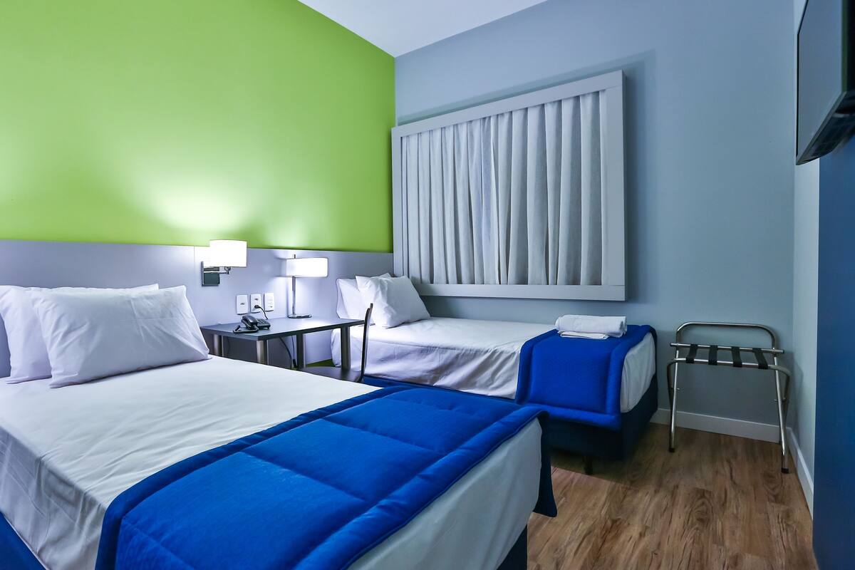 Quarto de hotel com duas camas, uma escrivaninha, uma cadeira, um abajur, um radiador e uma janela grande com cortinas.