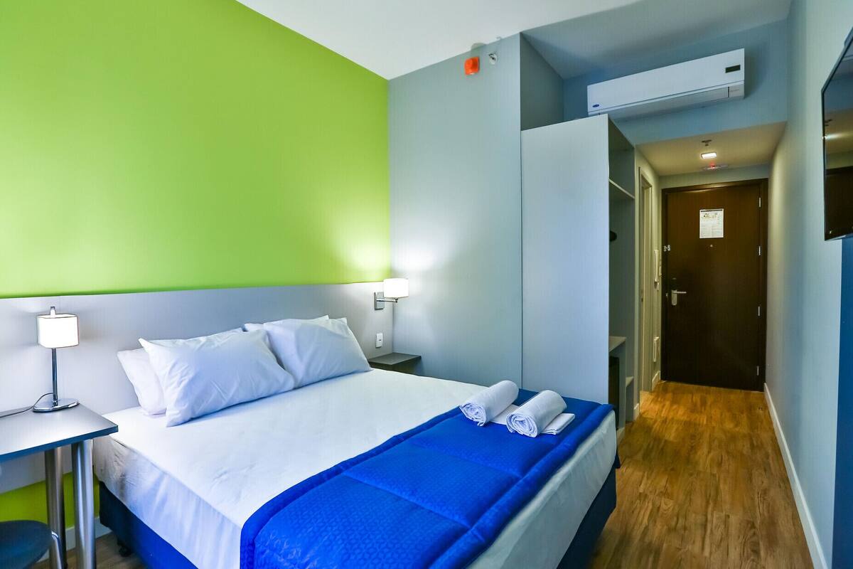 Quarto de hotel com cama, mesa de cabeceira, abajur e aparelho de ar condicionado.