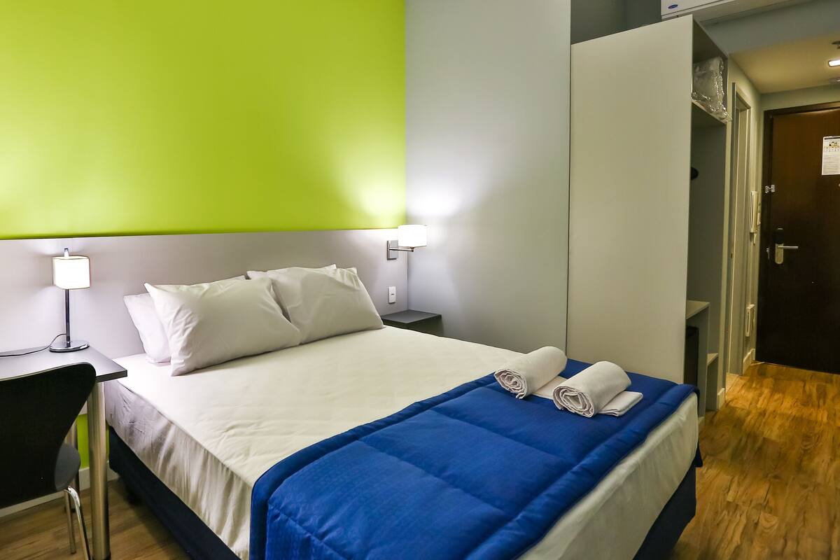 Quarto de hotel com cama, mesas de cabeceira, escrivaninha e guarda-roupa.