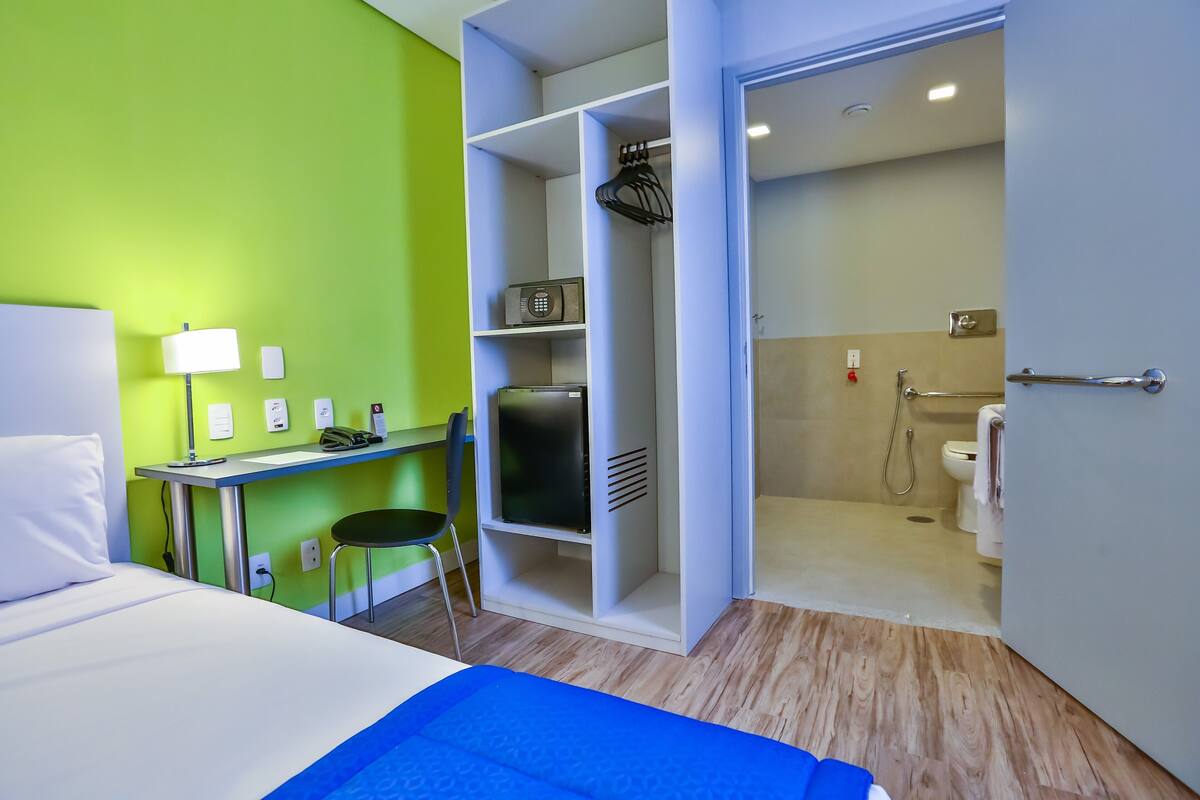 Quarto de hotel com cama, escrivaninha com luminária, cadeira, guarda-roupa e chuveiro.