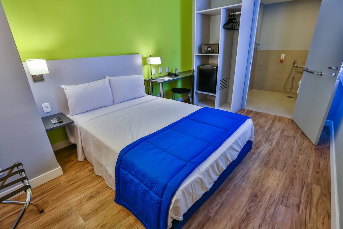 Quarto de hotel com cama, mesas de cabeceira, escrivaninha, área de chuveiro e armário.