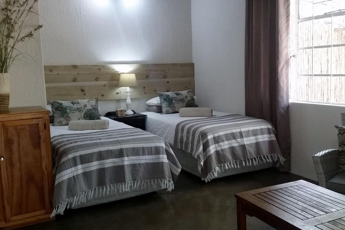 Quarto com duas camas, uma cabeceira de madeira, mesa de cabeceira com abajur, uma cadeira e uma janela com cortinas.