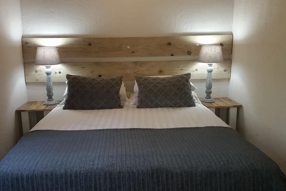 Uma cama com cabeceira de madeira, duas mesas de cabeceira com abajures e um cobreleito cinza.