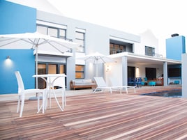Uma área externa moderna com piscina, deck de madeira, cadeiras de estar brancas e guarda-sóis.