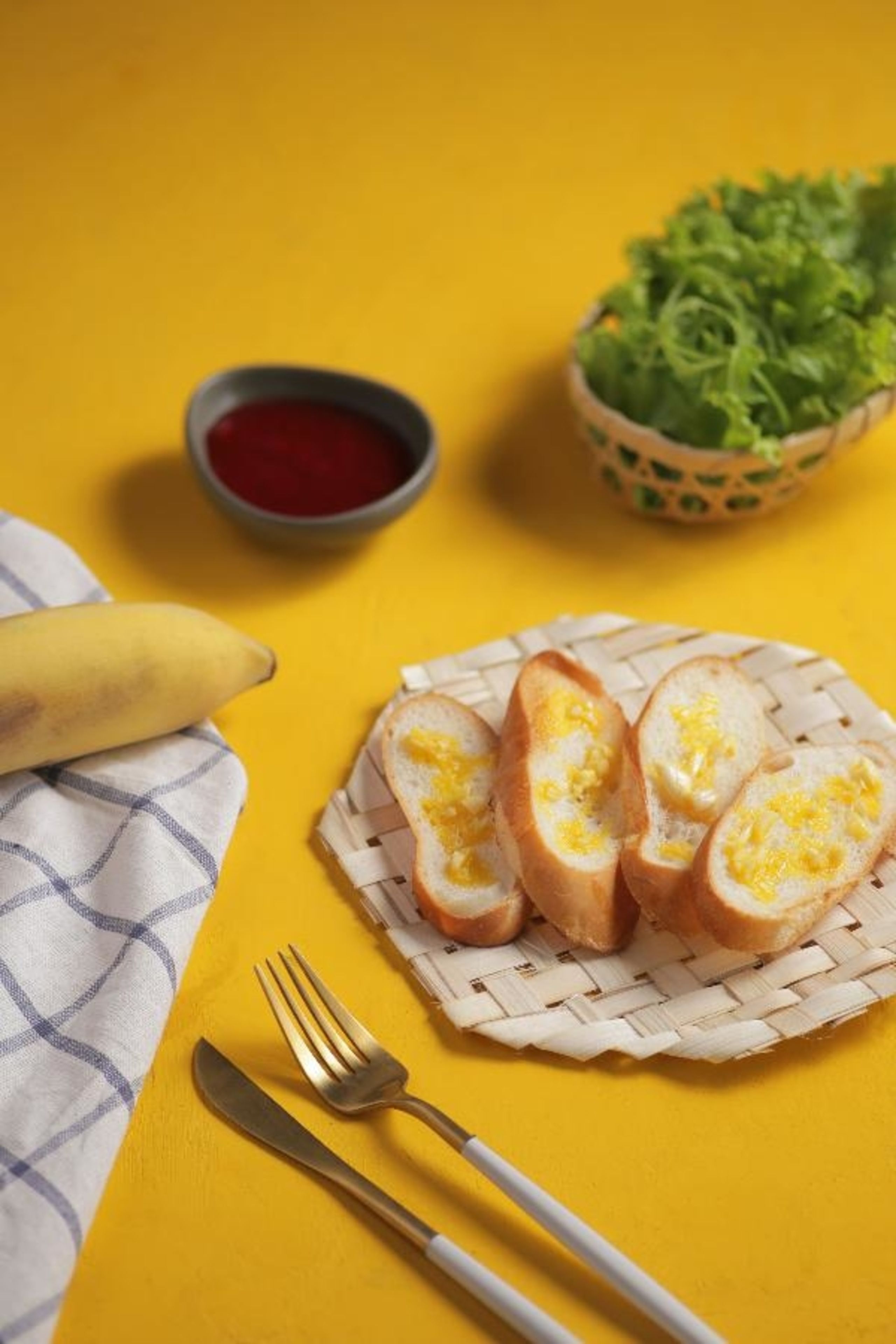 Um prato com pão e ovo, uma banana, um garfo, uma faca e uma pequena tigela de molho vermelho sobre uma mesa amarela.