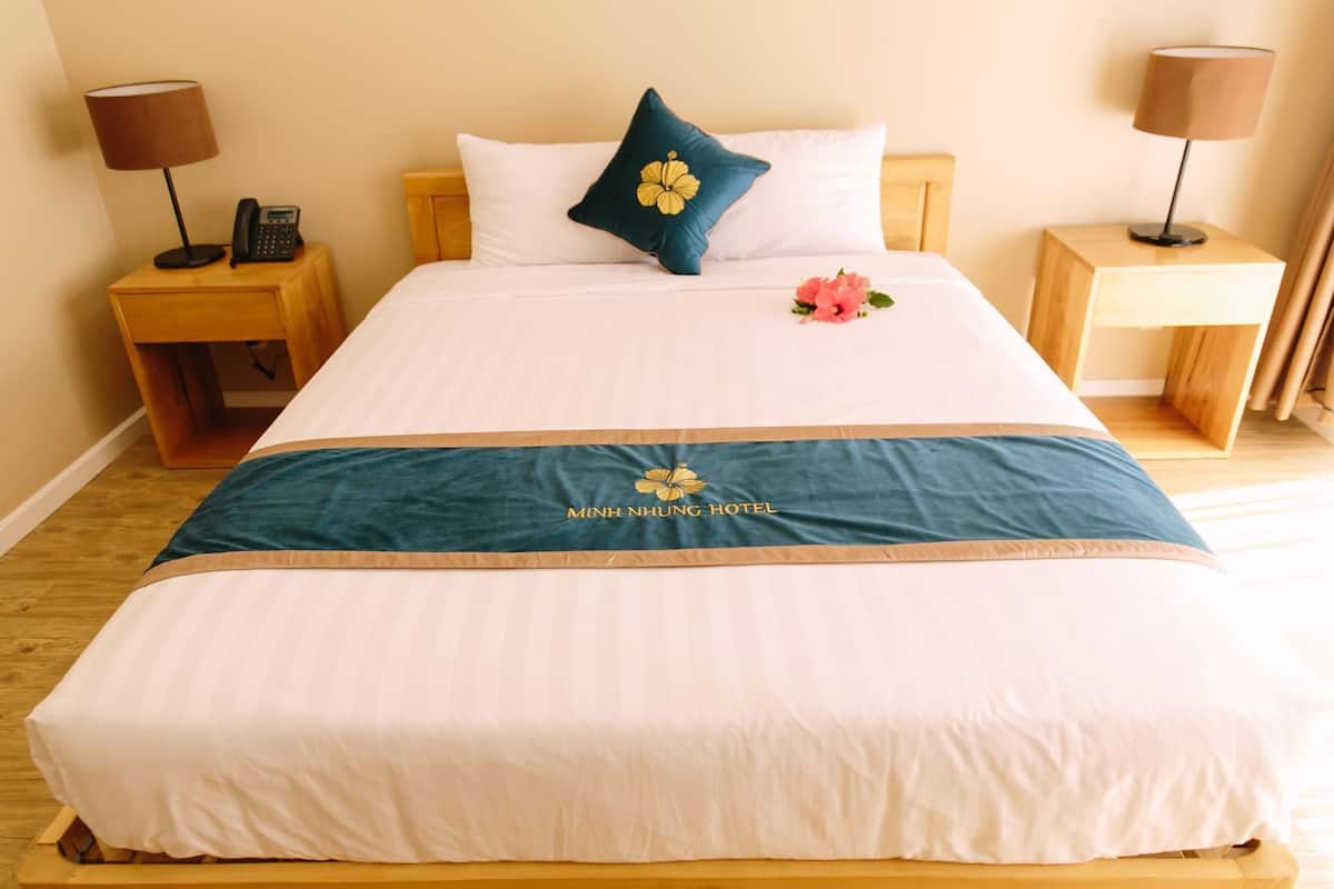 Quarto de hotel com cama, duas mesas de cabeceira, um criado-mudo, telefone e almofada decorativa.