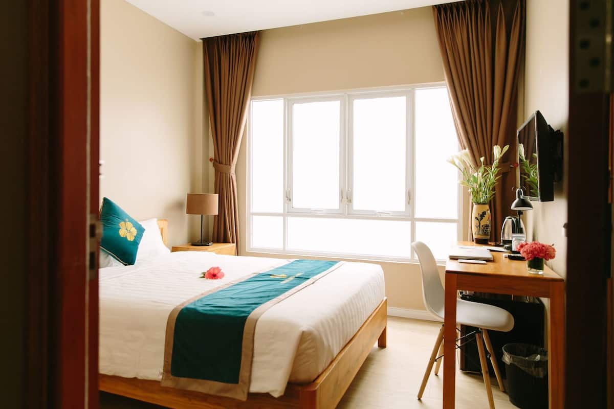 Quarto de hotel com cama, escrivaninha, cadeira e janela com cortinas.