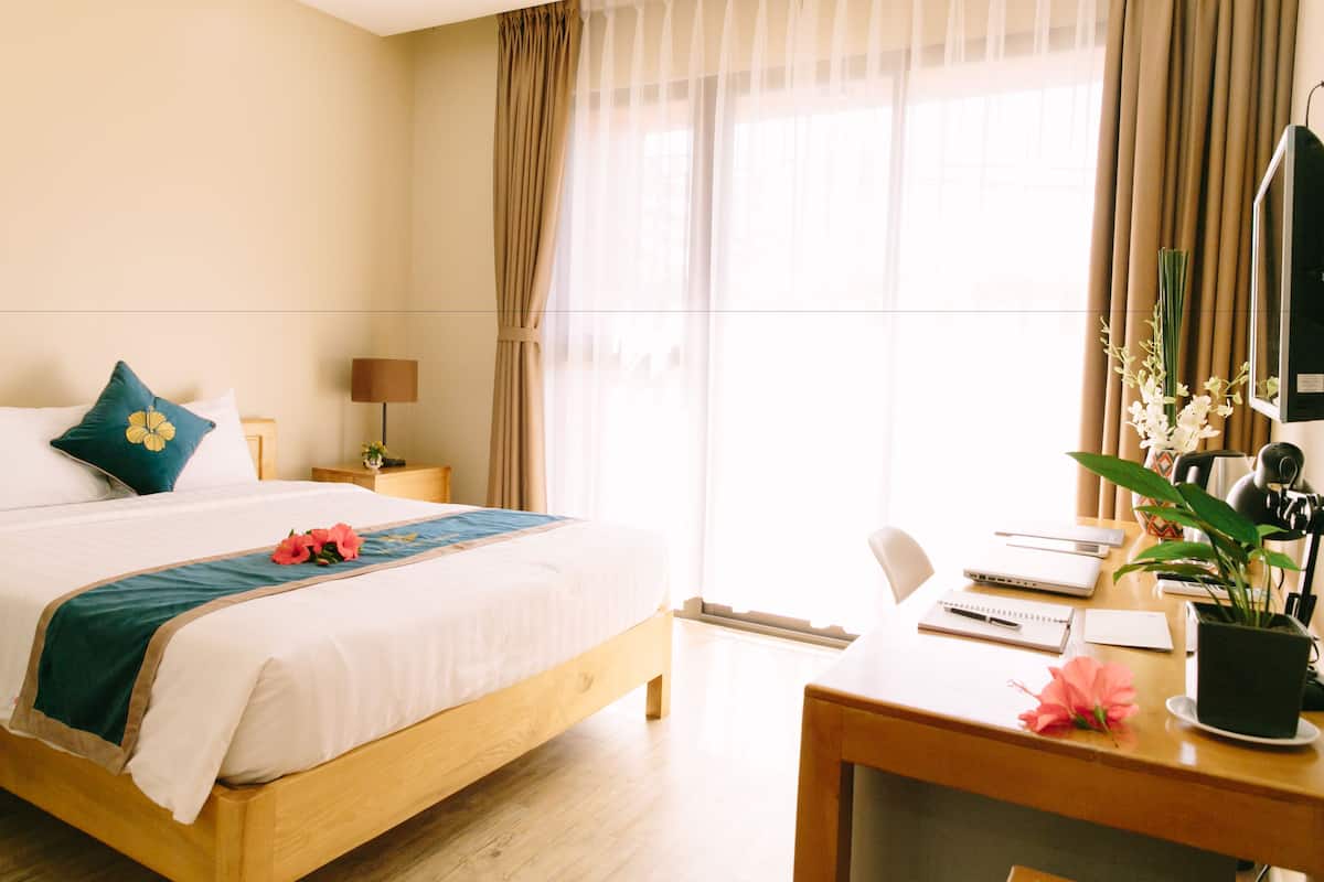 Quarto de hotel com cama, escrivaninha, luminária, planta em vaso e janela com cortinas.