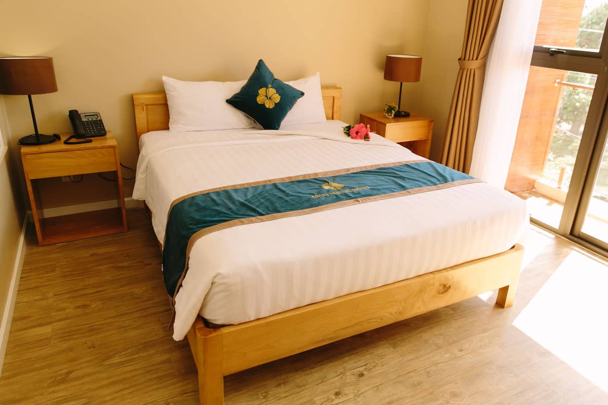 Cama bem arrumada com estrutura de madeira e edredom branco e azul-esverdeado. Há duas mesas de cabeceira, uma com luminárias e telefone.