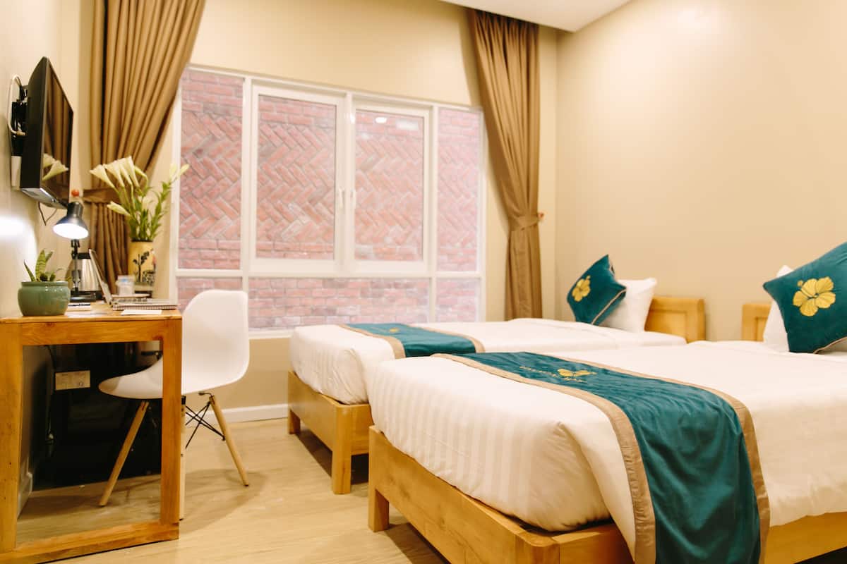 Quarto de hotel com duas camas de madeira, uma escrivaninha com cadeira, uma luminária de mesa e uma janela com cortinas.