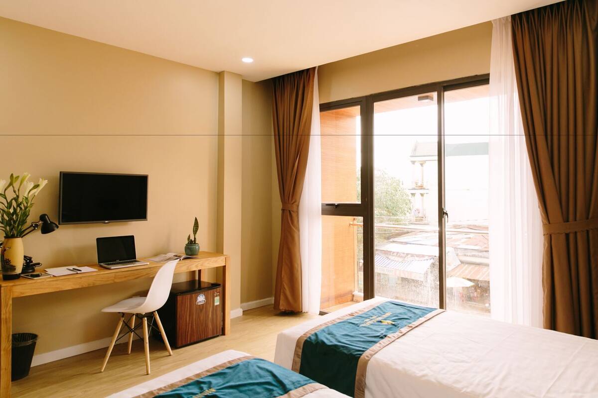 Quarto de hotel com escrivaninha, cadeira, televisão, duas camas, uma pequena lixeira e uma porta de vidro corrediça para área externa.