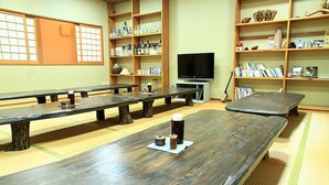 Um quarto japonês tradicional com esteiras de tatame, uma longa mesa de madeira, bancos, uma televisão e prateleiras com diversos objetos.