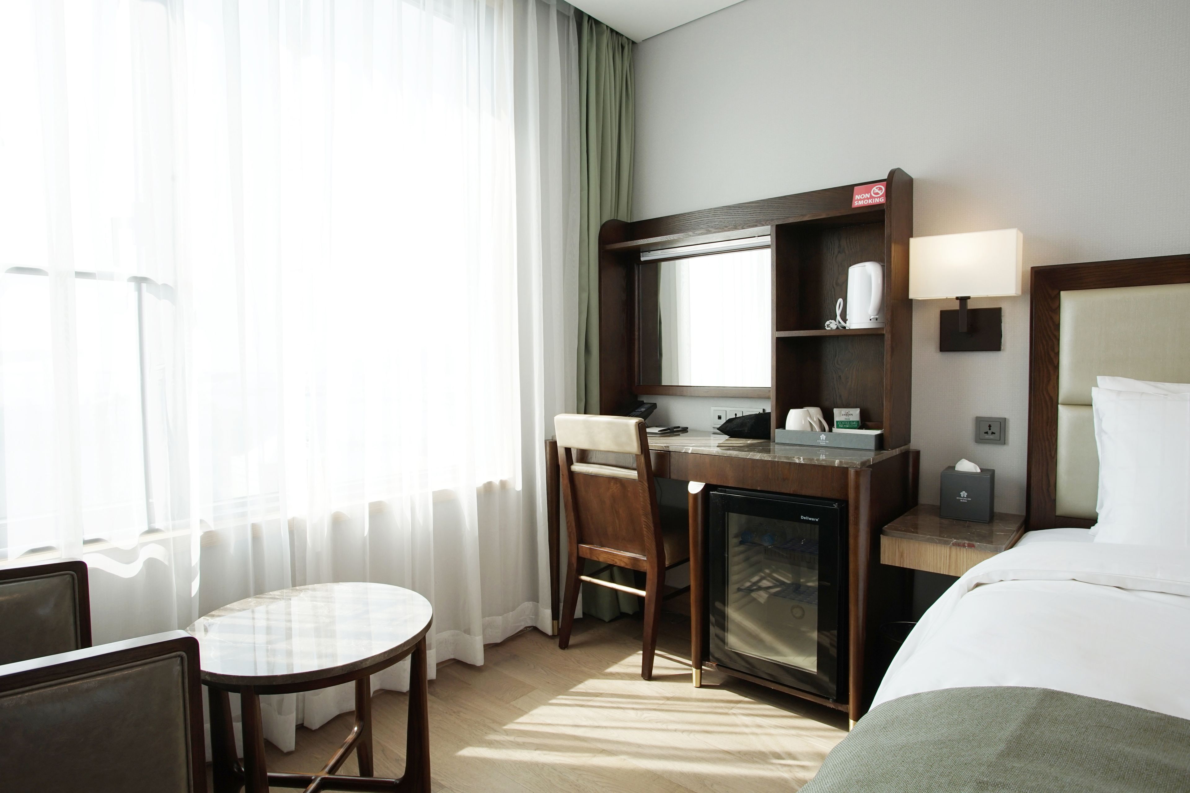 Quarto de hotel com cama, escrivaninha com cadeira, mesa pequena, minibar e janela com cortinas.
