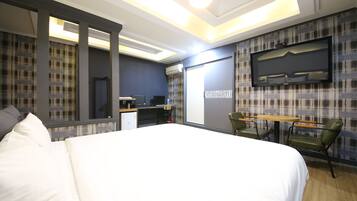 Quarto de hotel com uma cama grande, uma escrivaninha com computador, uma TV fixada na parede e um espaço de estar com cadeiras.