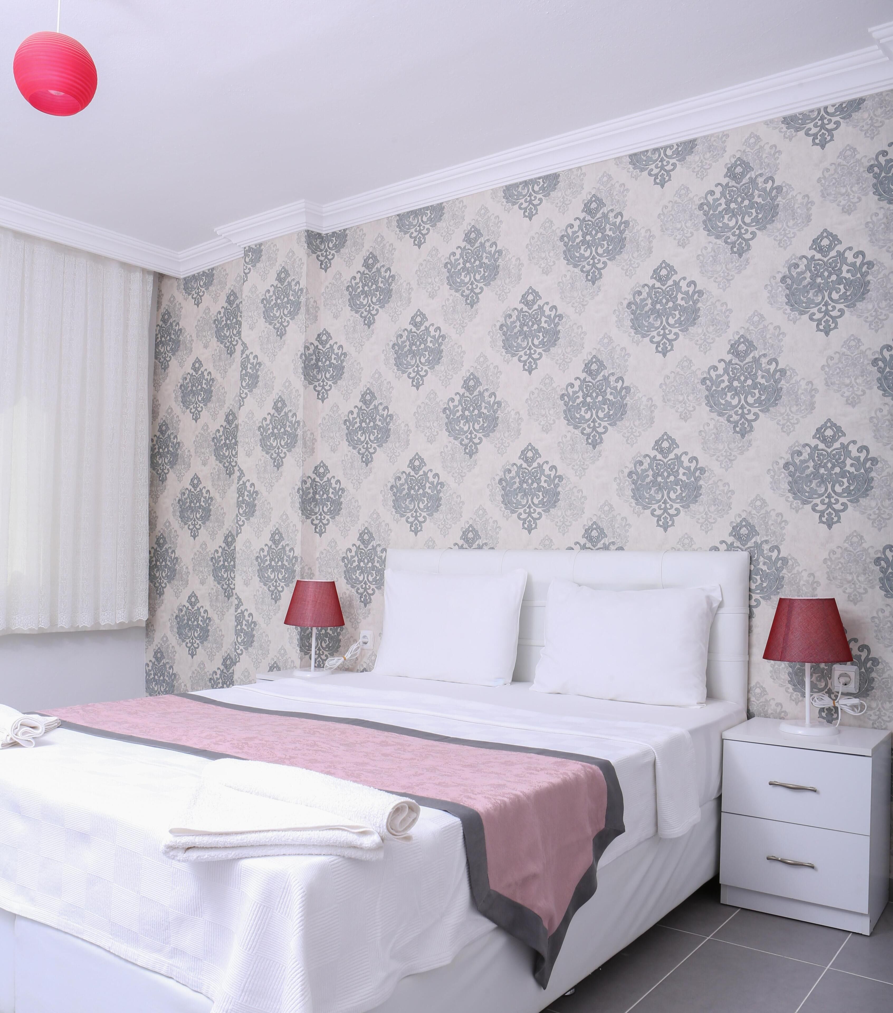 Cama bem arrumada com manta branca e rosa, mesa de cabeceira com abajur e papel de parede estampado.