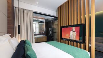 Quarto de hotel moderno com uma cama grande, uma televisão fixada em um painel de madeira e uma porta de vidro deslizante que dá para o banheiro.