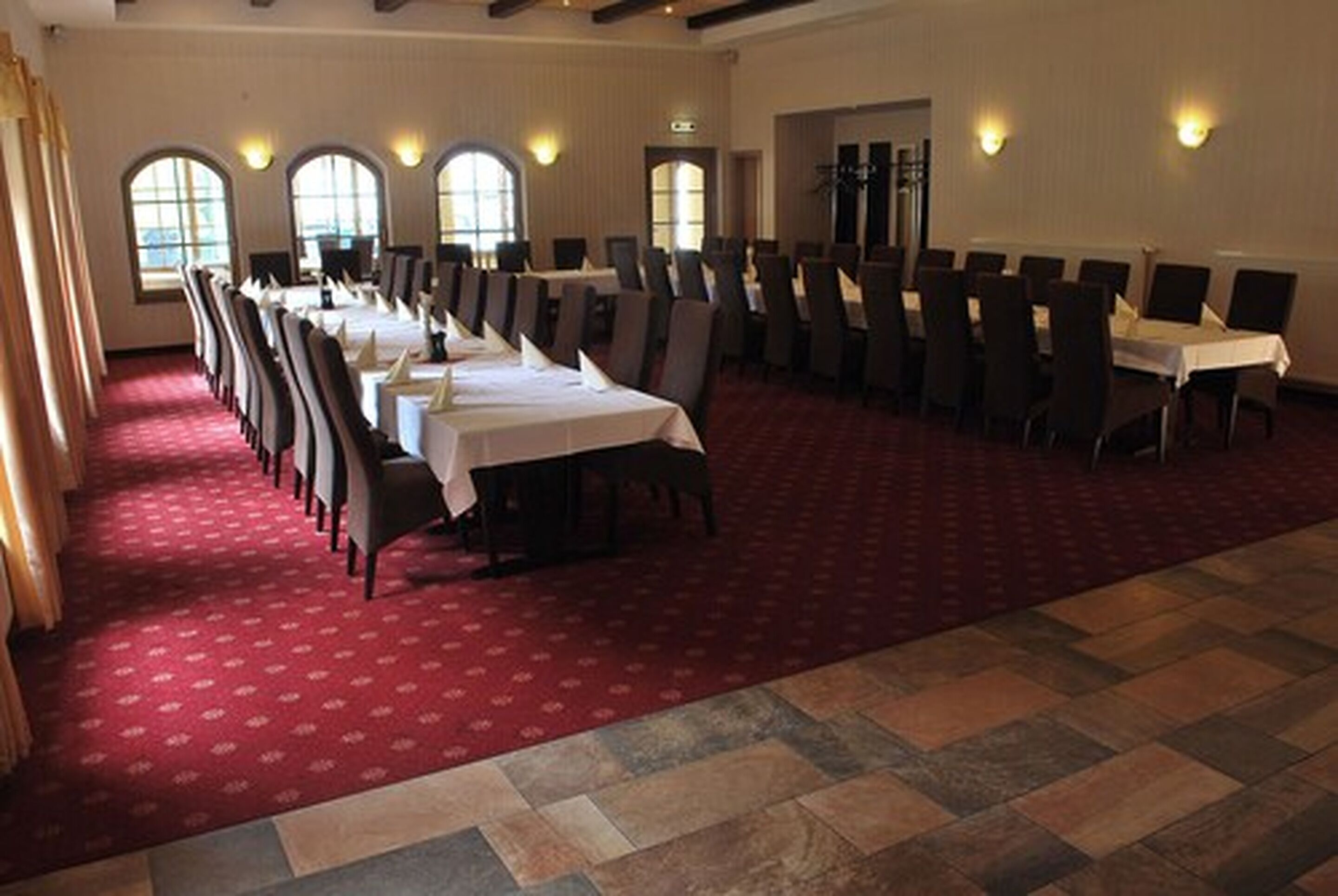 Uma sala de banquetes com mesas e cadeiras compridas, piso coberto de carpete e janelas amplas.
