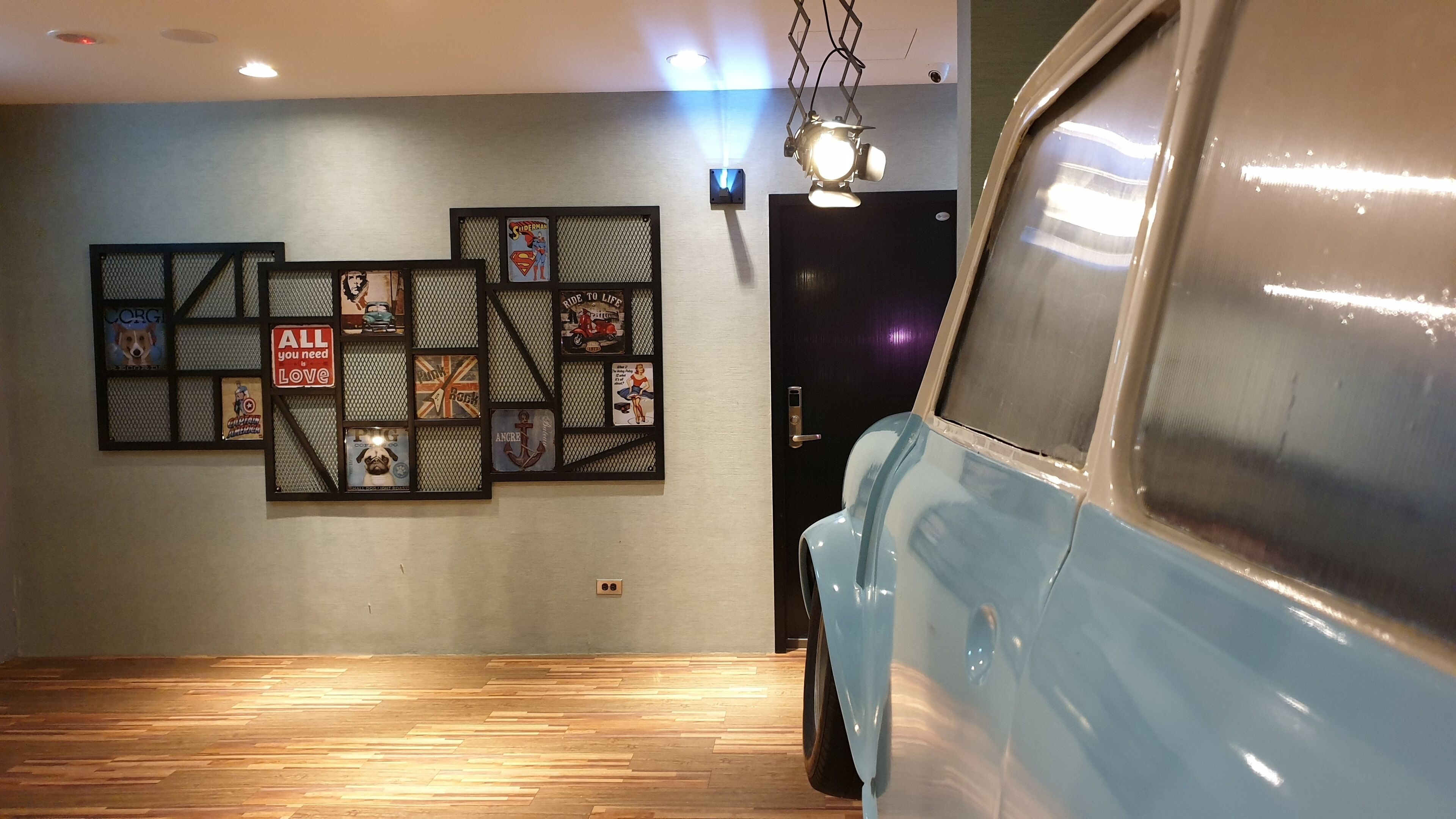 Uma garagem com um carro antigo, piso de madeira e uma parede decorada com diversos cartazes e placas.