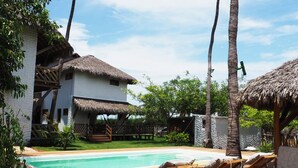 Uma área junto à piscina com espreguiçadeiras, um edifício com telhado de palha e palmeiras.