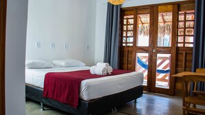 Quarto de hotel com uma cama, uma escrivaninha de madeira, um candeeiro pendente e vista para o exterior através de uma porta de madeira.