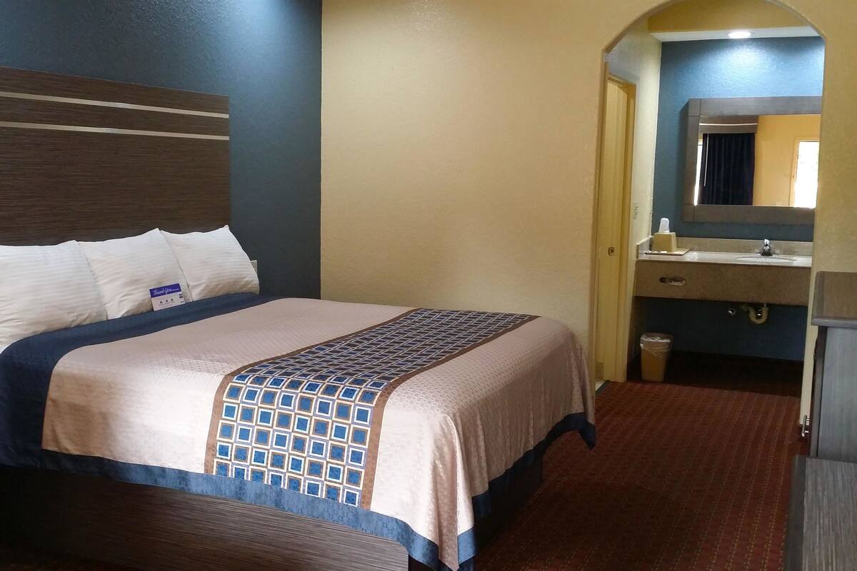 Quarto de hotel com cama, mesa de cabeceira, escrivaninha, banheiro com pia e espelho, e piso coberto por carpete.
