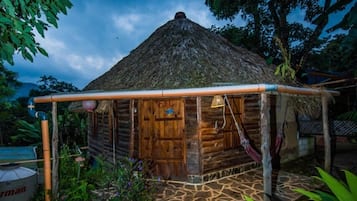 Uma cabana de madeira rústica com telhado de palha, rodeada por vegetação exuberante e um caminho de pedra.