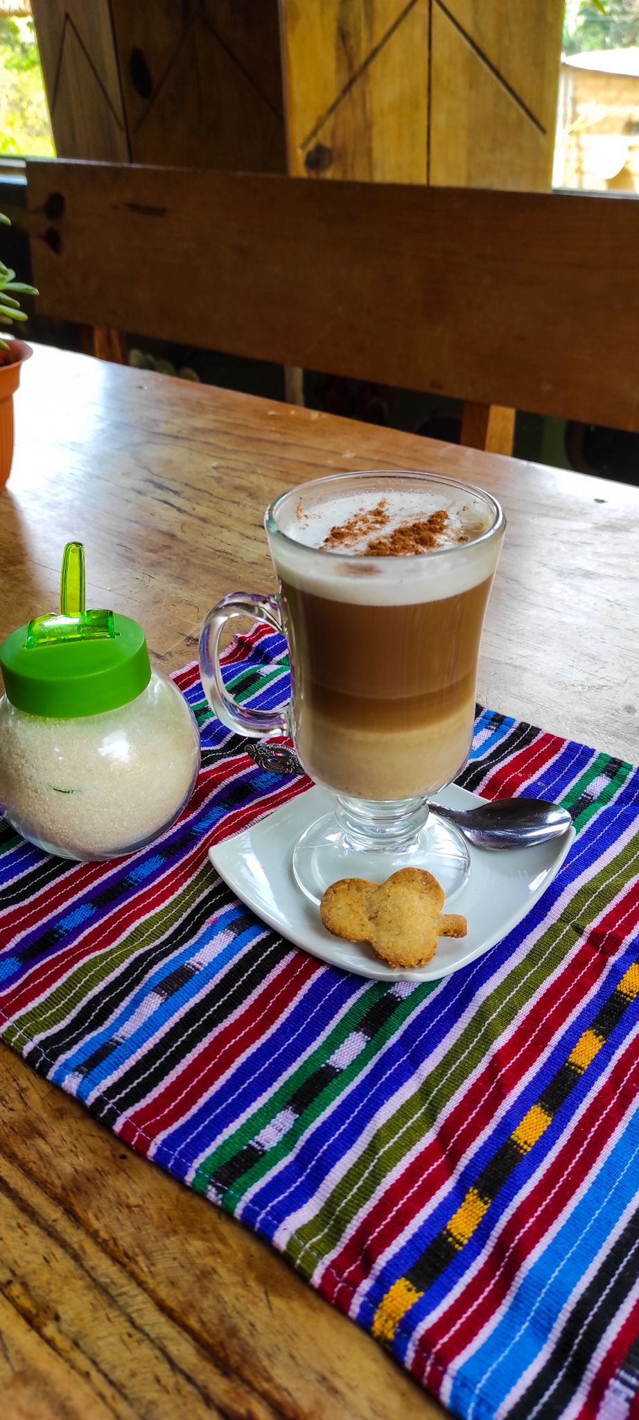 Uma chávena de café com uma colher sobre uma toalha de mesa colorida listrada, um pequeno frasco com tampa verde e um bolinho num prato branco.