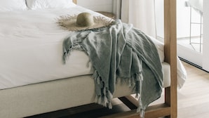 Quarto com cama de dossel de madeira, roupa de cama branca, chapéu de palha e manta cinza.